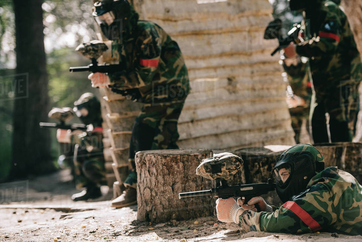Focused paintball player laying on ground and aiming with marker gun ...