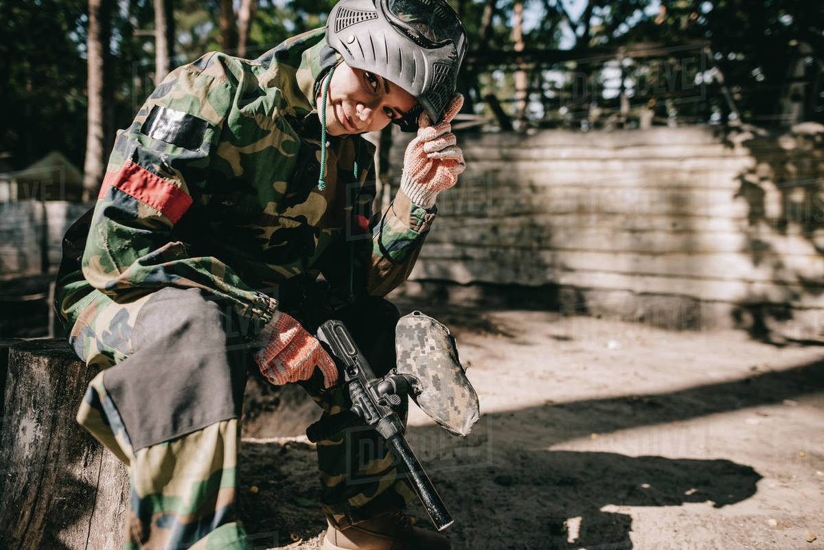 Smiling young female paintballer in camouflage uniform sitting with ...