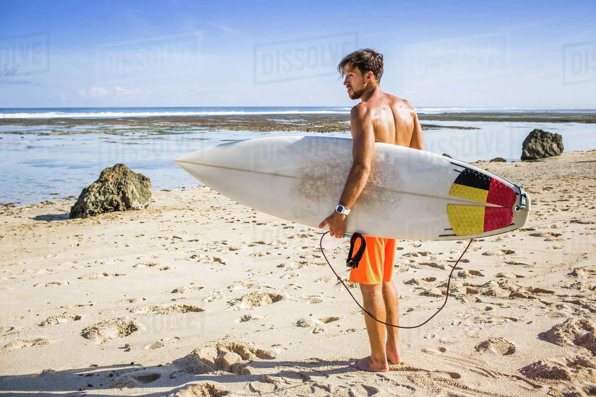 Side view of young surfer with surfing board standing on sandy beach on ...
