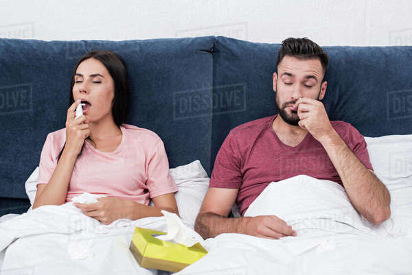 Sick couple using cough and nose spray while sitting in bed - Royalty ...