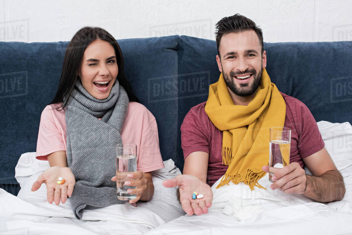Happy sick couple with pills and glasses of water sitting in bed and ...