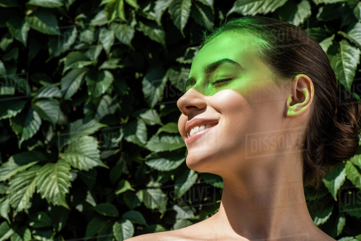 Young smiling woman with green shadow on face and leaves at background ...
