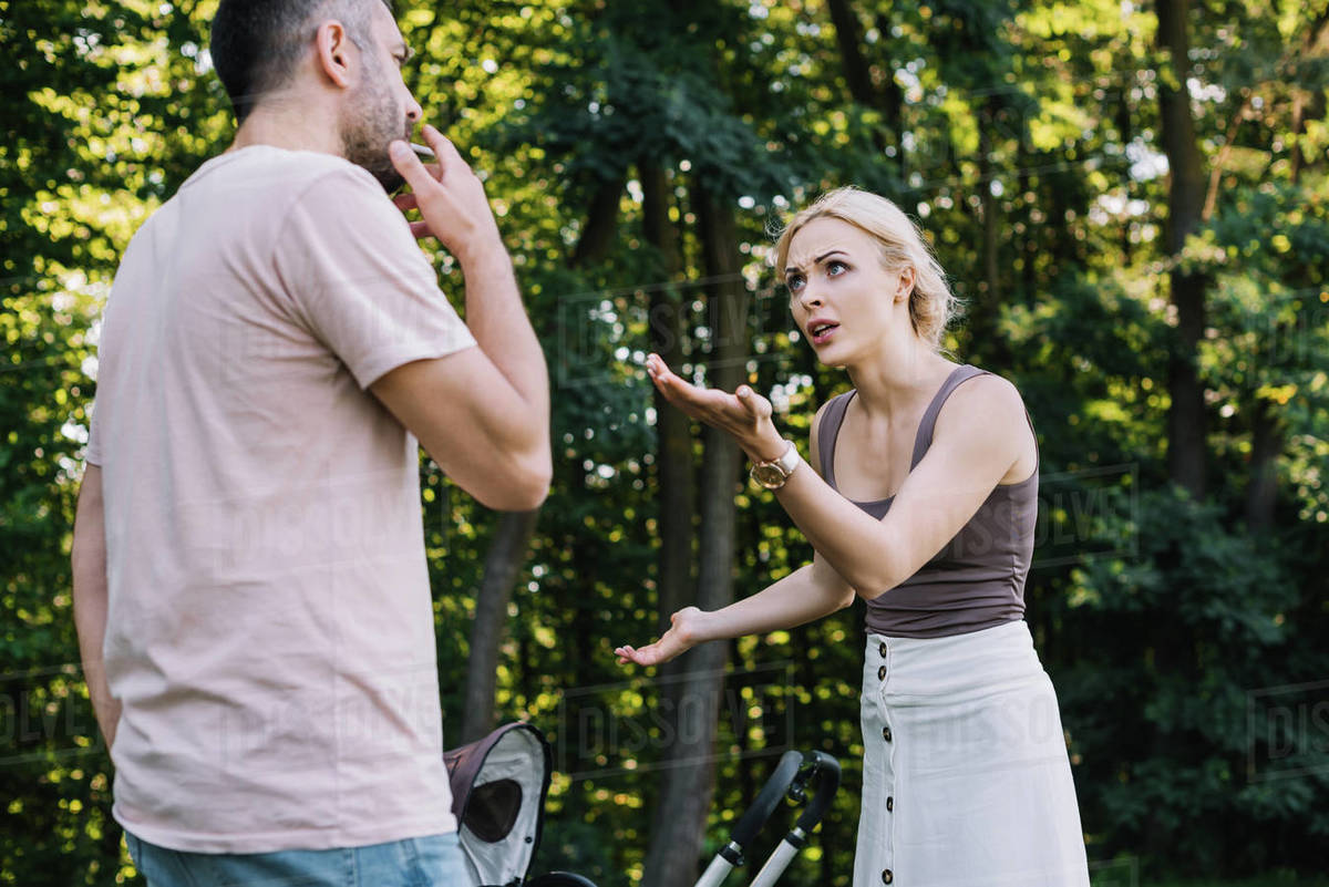 Father smoking cigarette near baby carriage in park, mother gesturing ...
