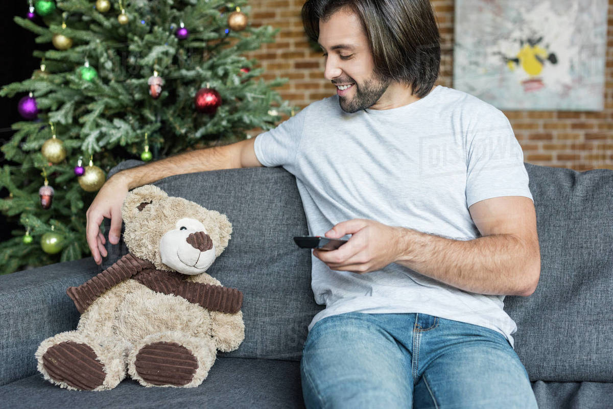 Smiling man holding remote controller and looking at teddy bear on ...