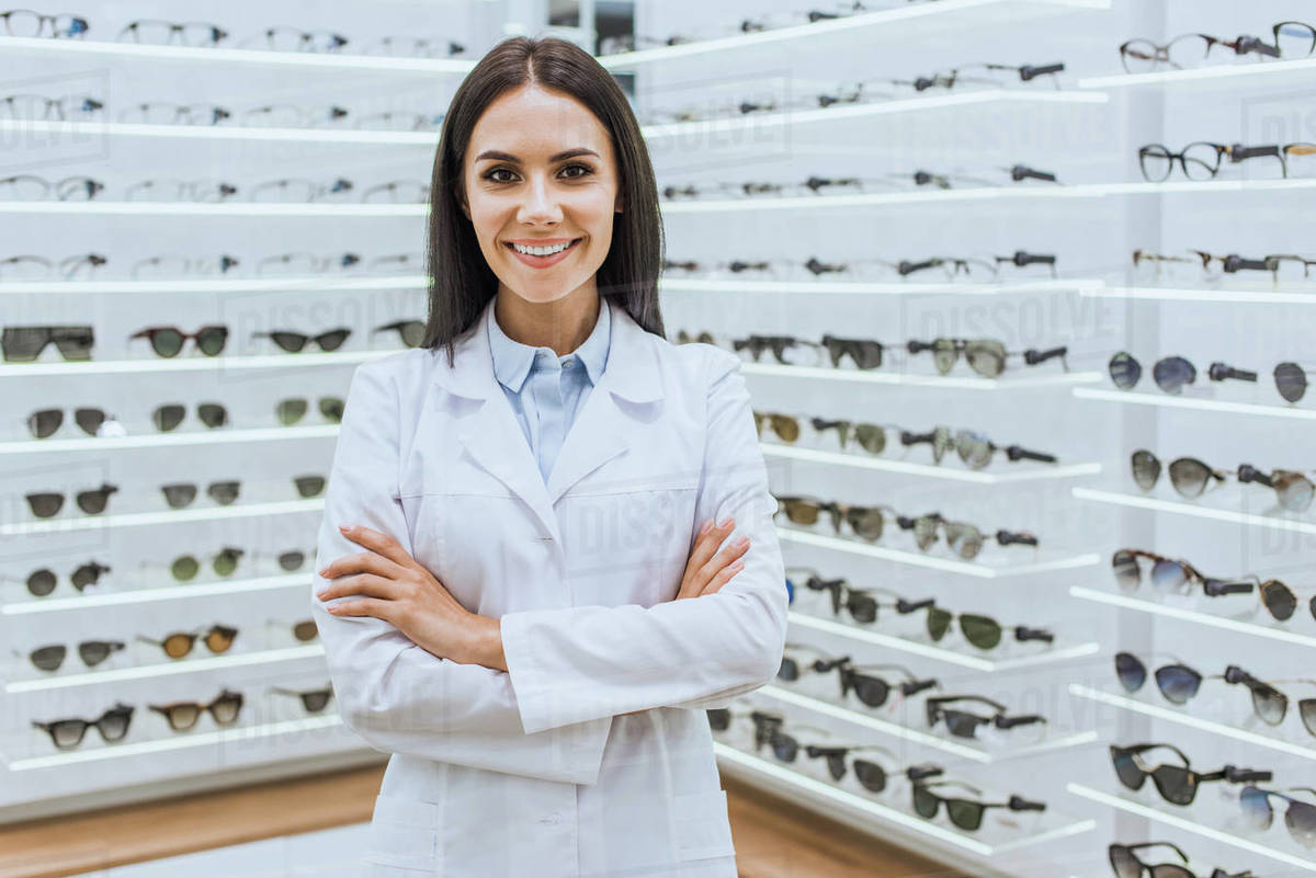 Professional smiling optometrist posing with crossed arms near shelves ...