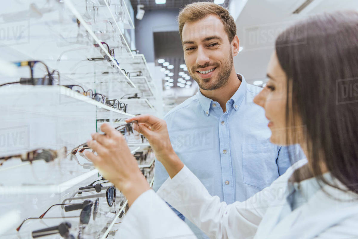 Side view of female oculist taking eyeglasses from shelves to happy man ...