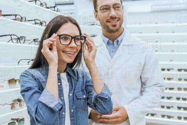 Happy woman choosing eyeglasses while male oculist standing near in ...