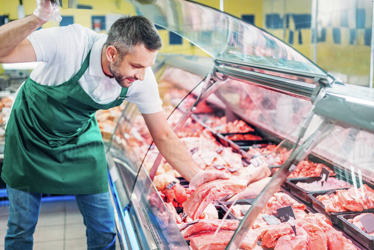 shop assistant in apron assorting fresh raw meat in supermarket ...