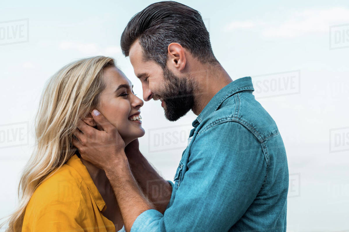 side view of smiling couple hugging and going to kiss against blue sky ...
