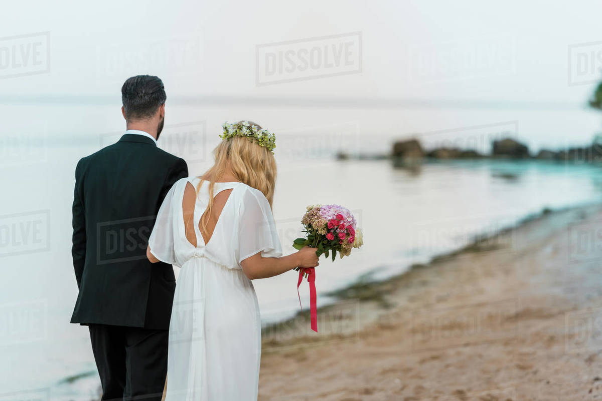 back view of wedding couple with bouquet walking on beach - Royalty ...
