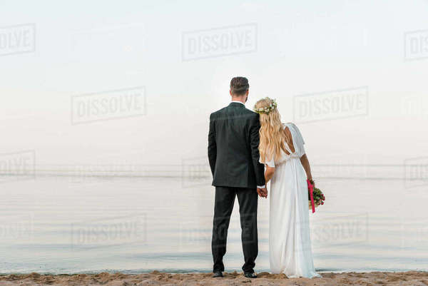 back view of wedding couple standing on beach with wedding bouquet and ...