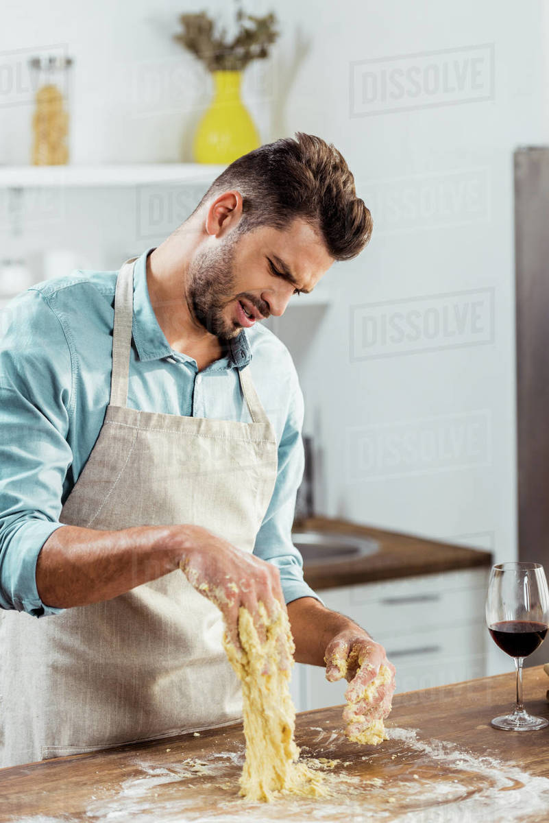Frustrated young man in apron looking at spoiled dough on hands in ...