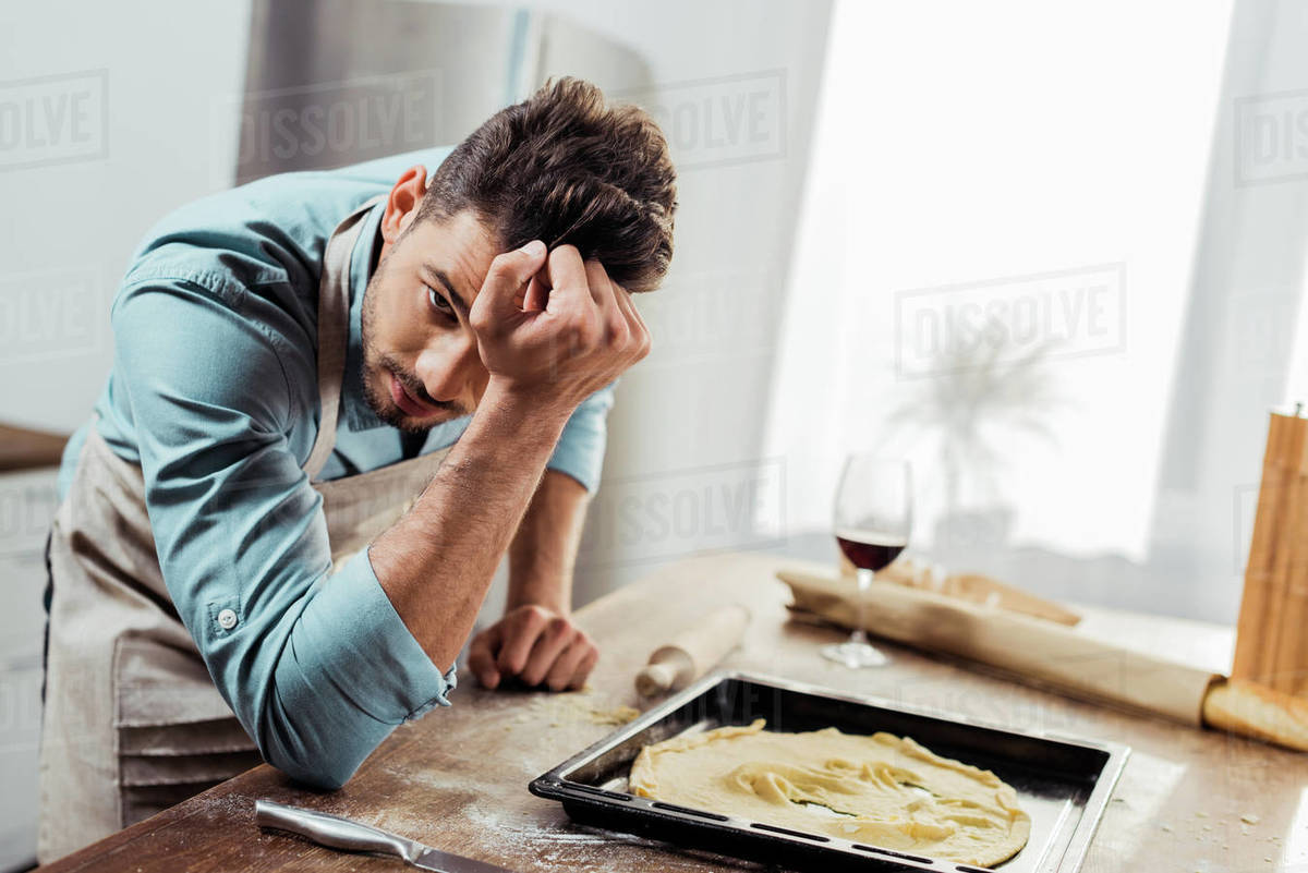 Upset young man in apron looking at camera while leaning at table with ...