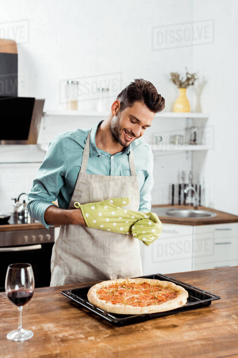 Smiling young man in apron and potholders looking at fresh homemade ...