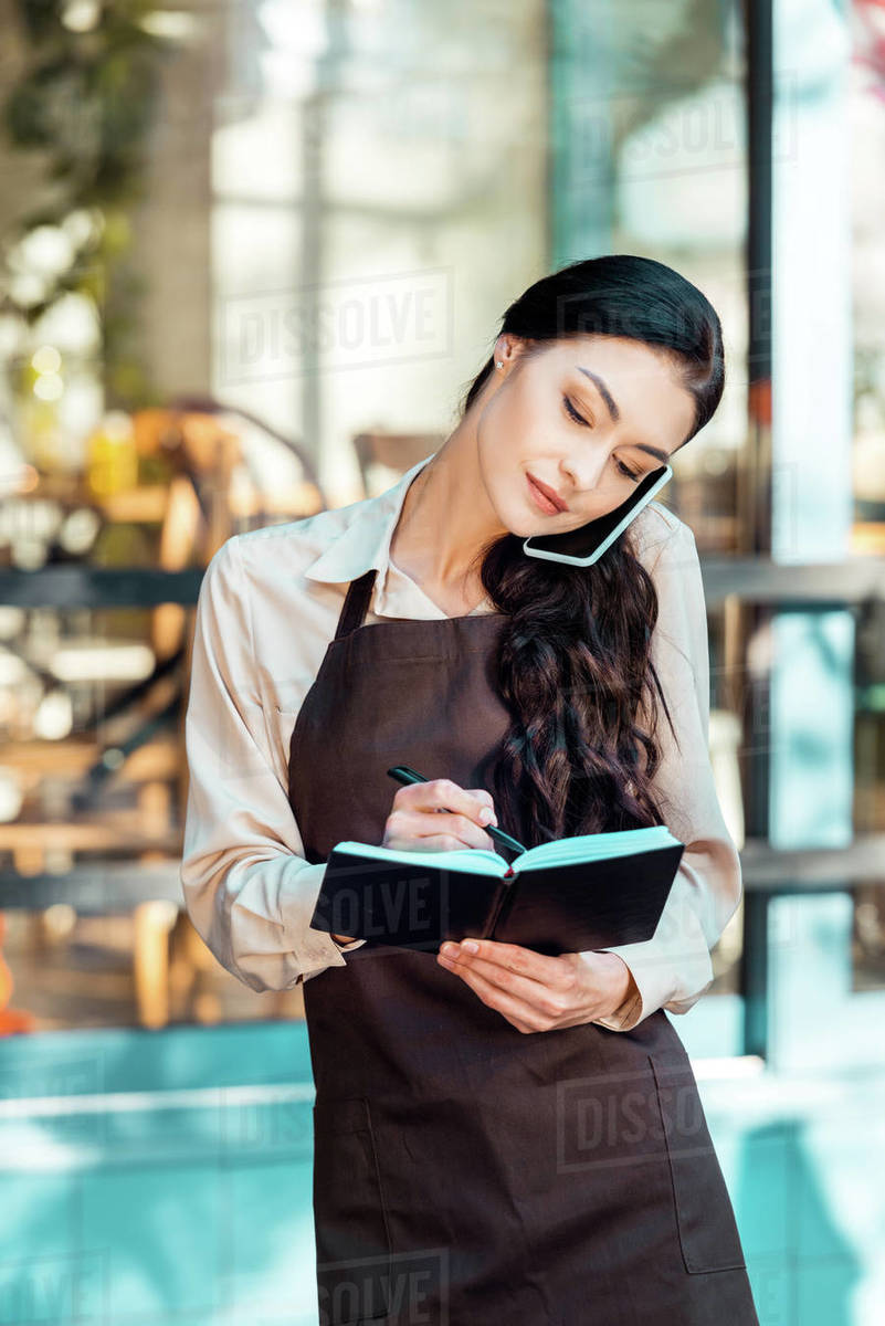 Beautiful waitress in apron talking by smartphone on street near cafe ...