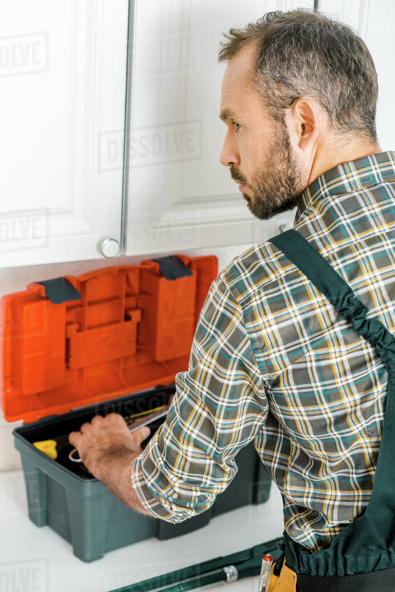 Side view of handsome plumber taking tools from toolbox in kitchen ...