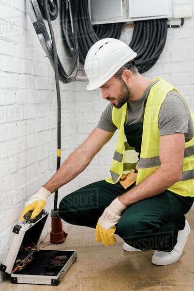 Handsome electrician squatting near electrical box and looking into ...