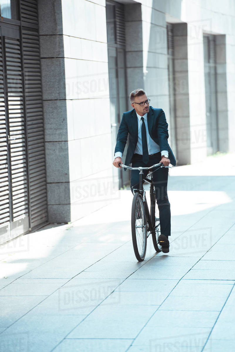 Handsome middle aged businessman in suit and eyeglasses riding bicycle ...