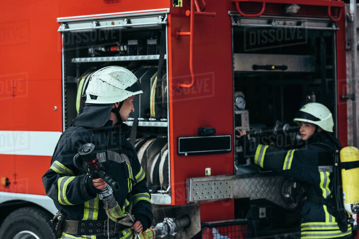 male firefighter in uniform holding water hose while colleague checking ...