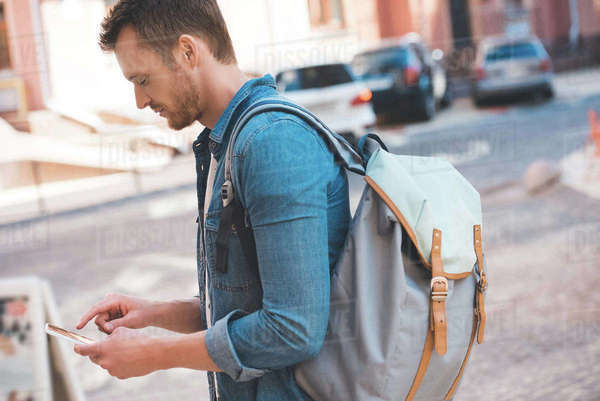 Handsome young man with backpack walking by street and using smartphone ...