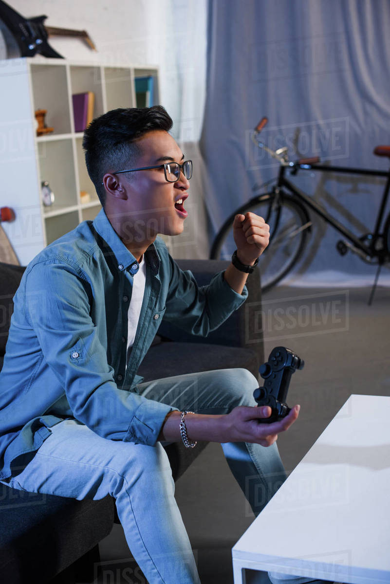 Excited young Asian man playing video game with joystick and win at ...