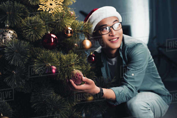 Young happy Asian man in Santa hat decorating Christmas tree and ...