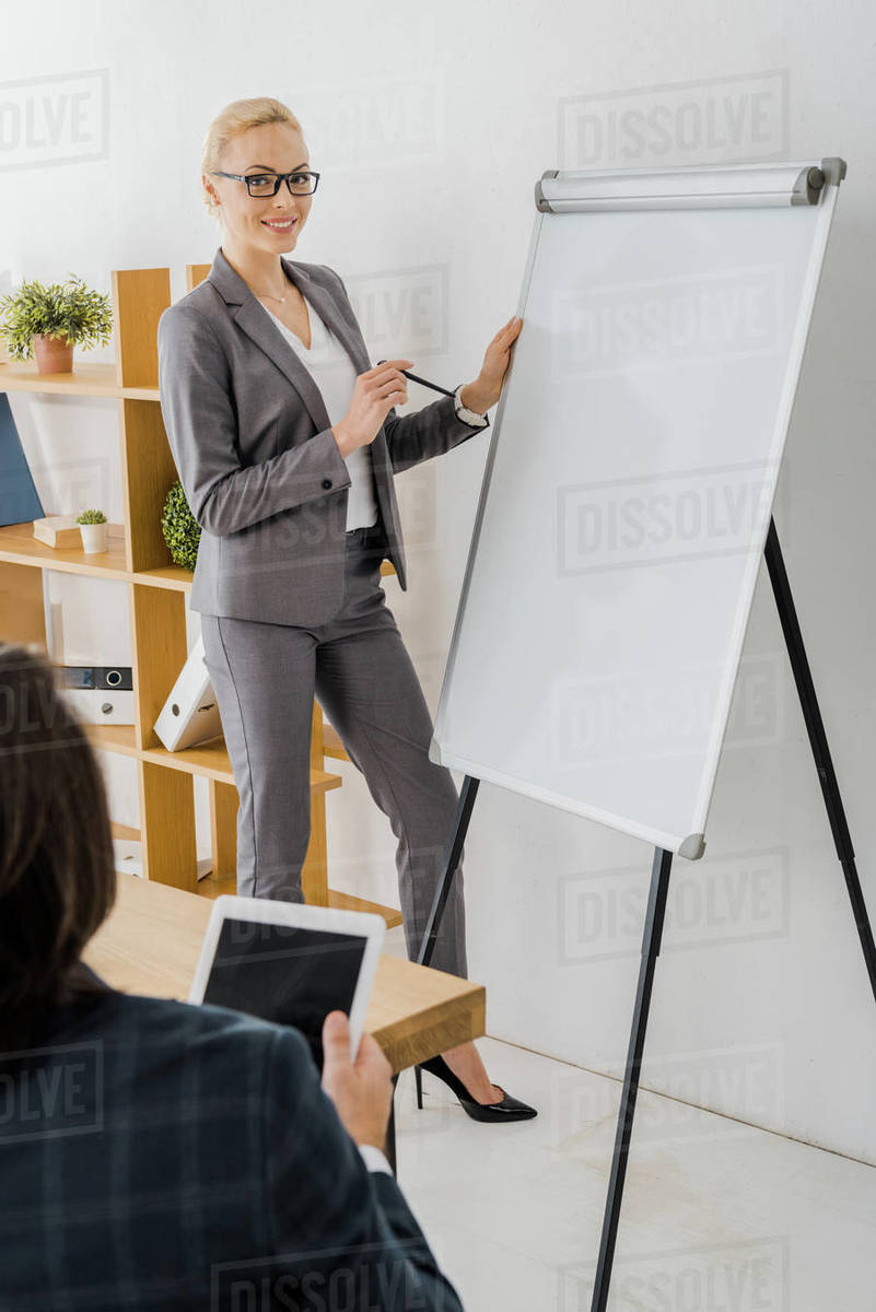 Young smiling woman pointing at white board and man sitting at table ...