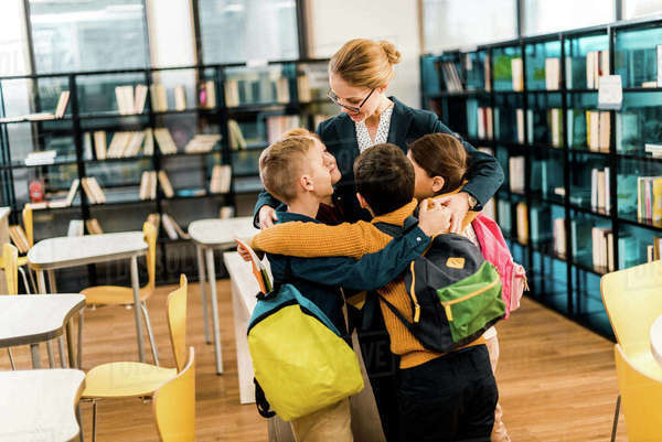 Happy schoolchildren with backpacks hugging librarian in library ...