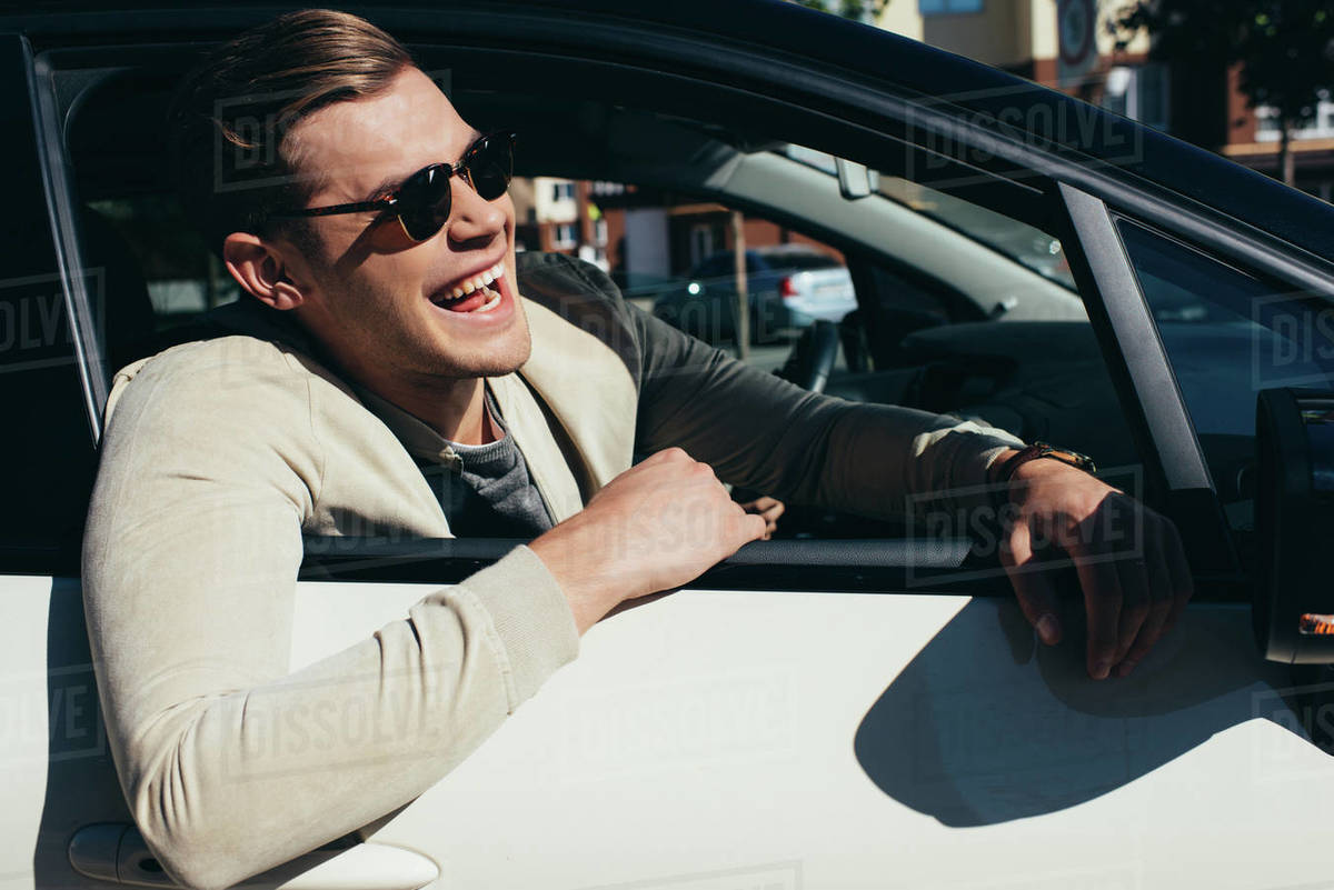 Side view of cheerful young man leaning out car window of driving ...