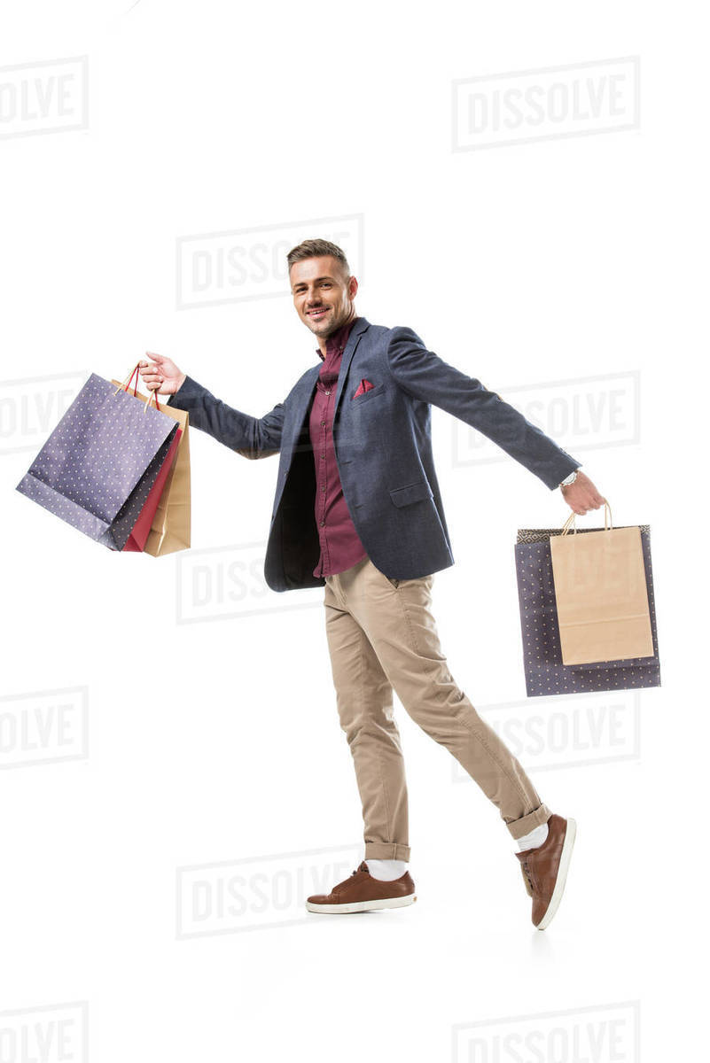 Smiling adult male shopper in jacket walking with colorful paper bags ...