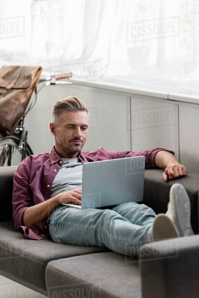 Handsome freelancer laying on sofa and working on laptop at home office ...