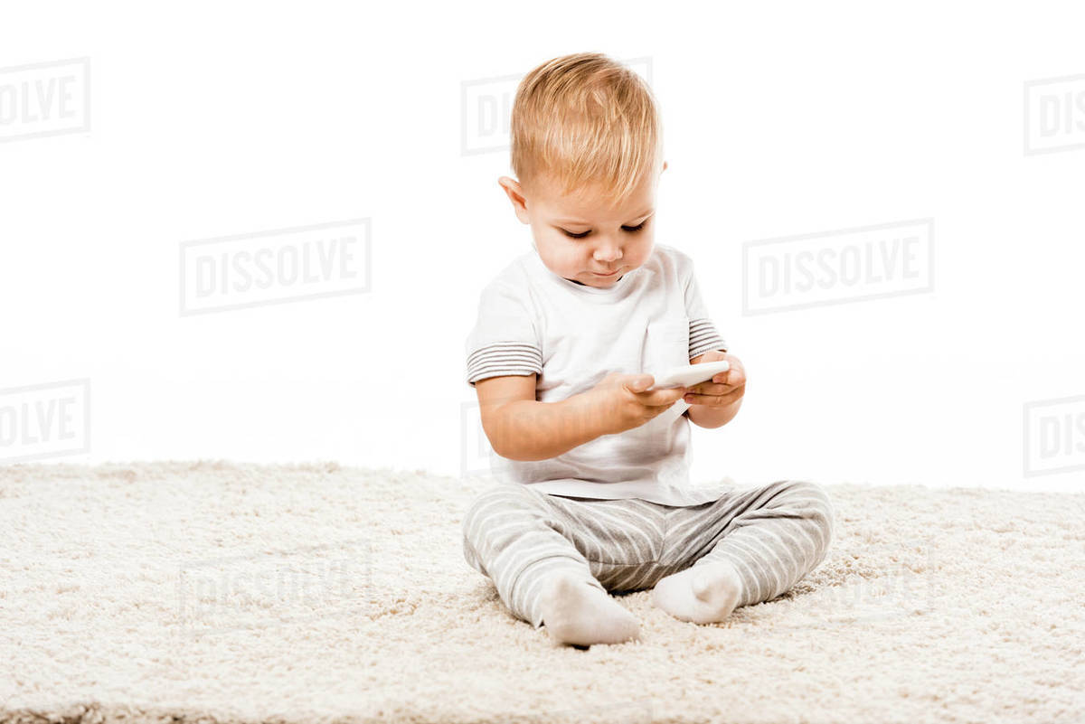 Adorable little boy with smartphone sitting on carpet isolated on white ...