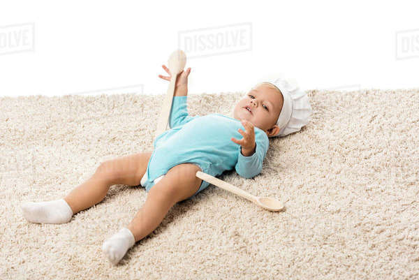 Toddler boy in chefs hat with two big wooden spoons lying on carpet and ...