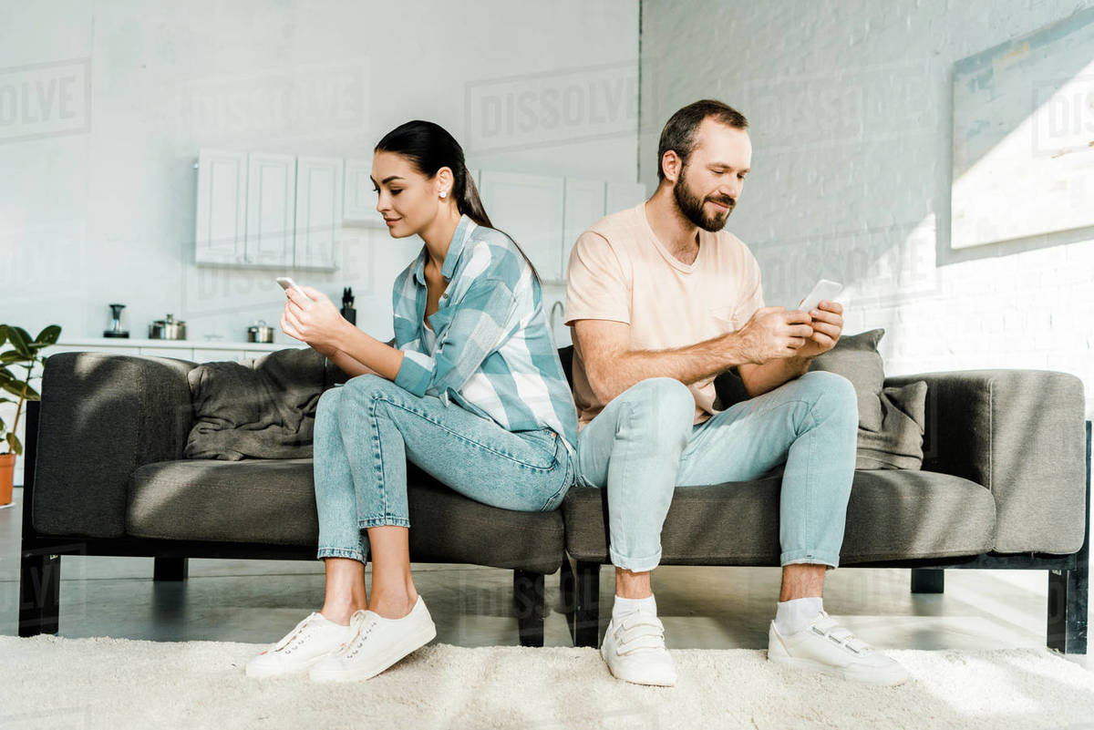 Couple sitting on couch, facing opposite direction and using ...