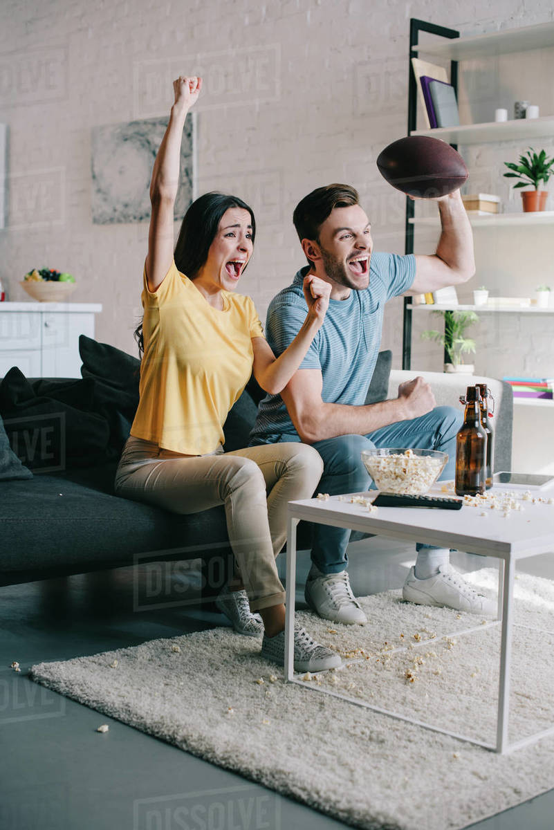 Emotional young couple cheering for American football game at home ...