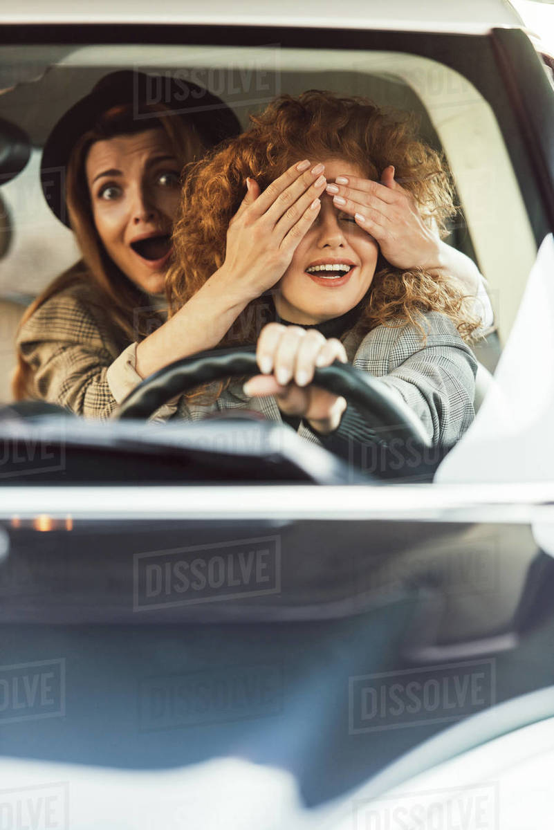 Shocked woman covering eyes to female friend while she driving car ...
