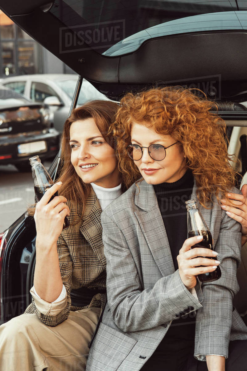 Stylish women in jackets looking away and posing with soda bottles in ...