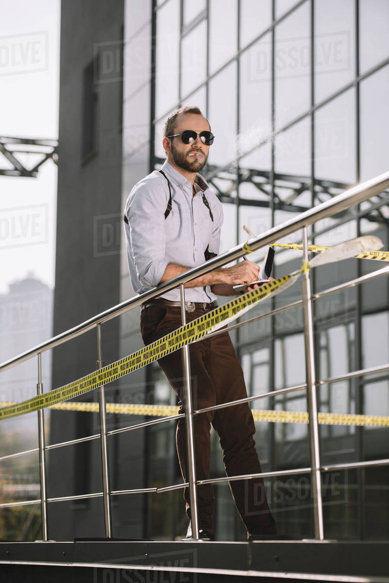 Male detective in sunglasses smoking and making notes leaning on the ...