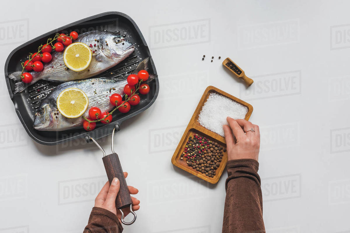 cropped image of woman taking salt at table with uncooked fish and ...