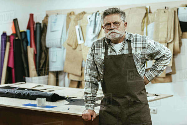 Confident male craftsman in apron and eyeglasses posing at studio ...