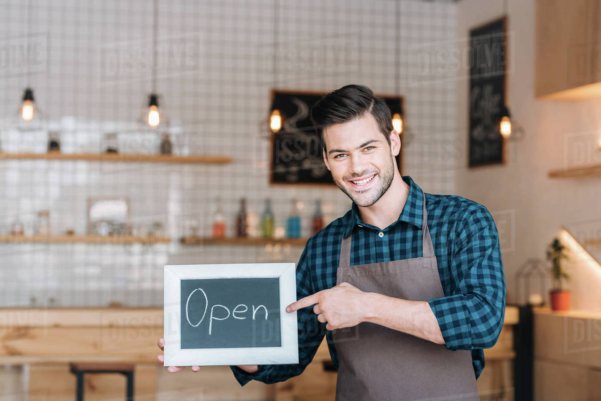 portrait of smiling barista pointing at chalkboard with open word in ...