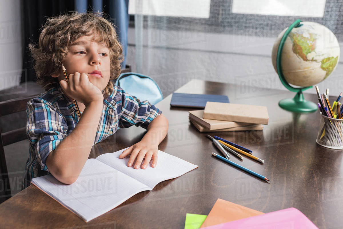 Thoughtful little boy looking away while doing homework at home ...