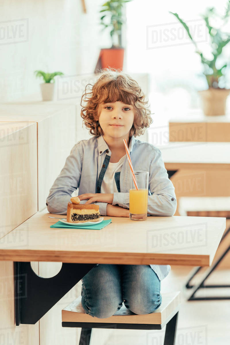 cute cheerful curly boy sitting in cafe in cafe with piece of cake and ...