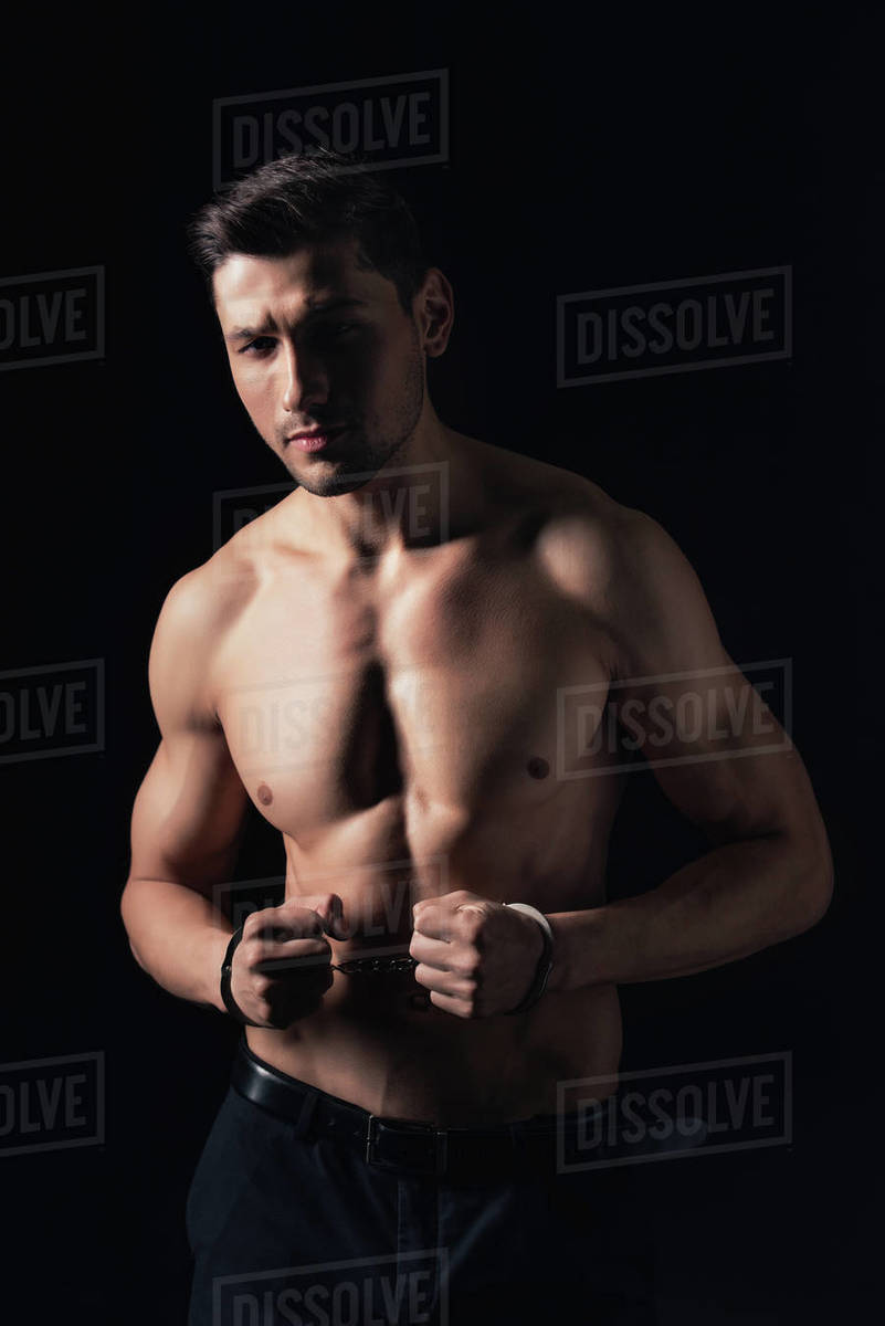 Handsome shirtless man in handcuffs looking at camera isolated on black ...