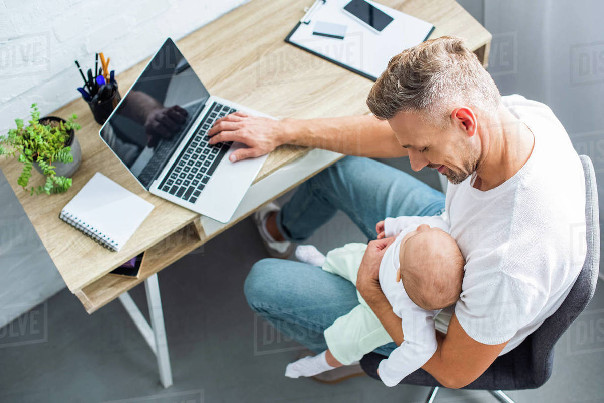 Father sitting at desk and using laptop with blank screen while holding ...