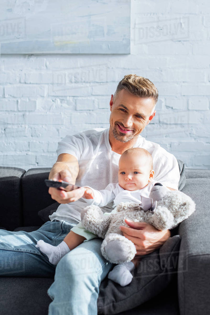 Happy father sitting on couch with baby daughter, holding remote ...