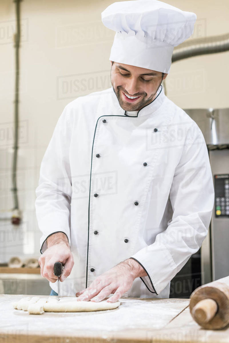 Handsome baker smiling and cutting uncooked dough on wooden table ...