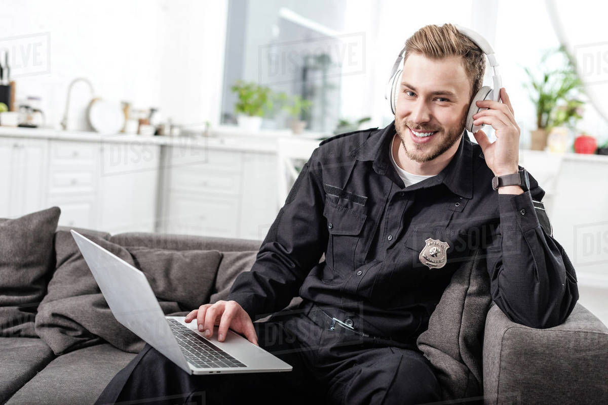 Handsome policeman sitting on sofa with laptop and listening to music ...