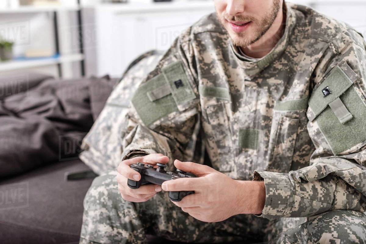 cropped view of army soldier sitting on couch at home and playing video ...