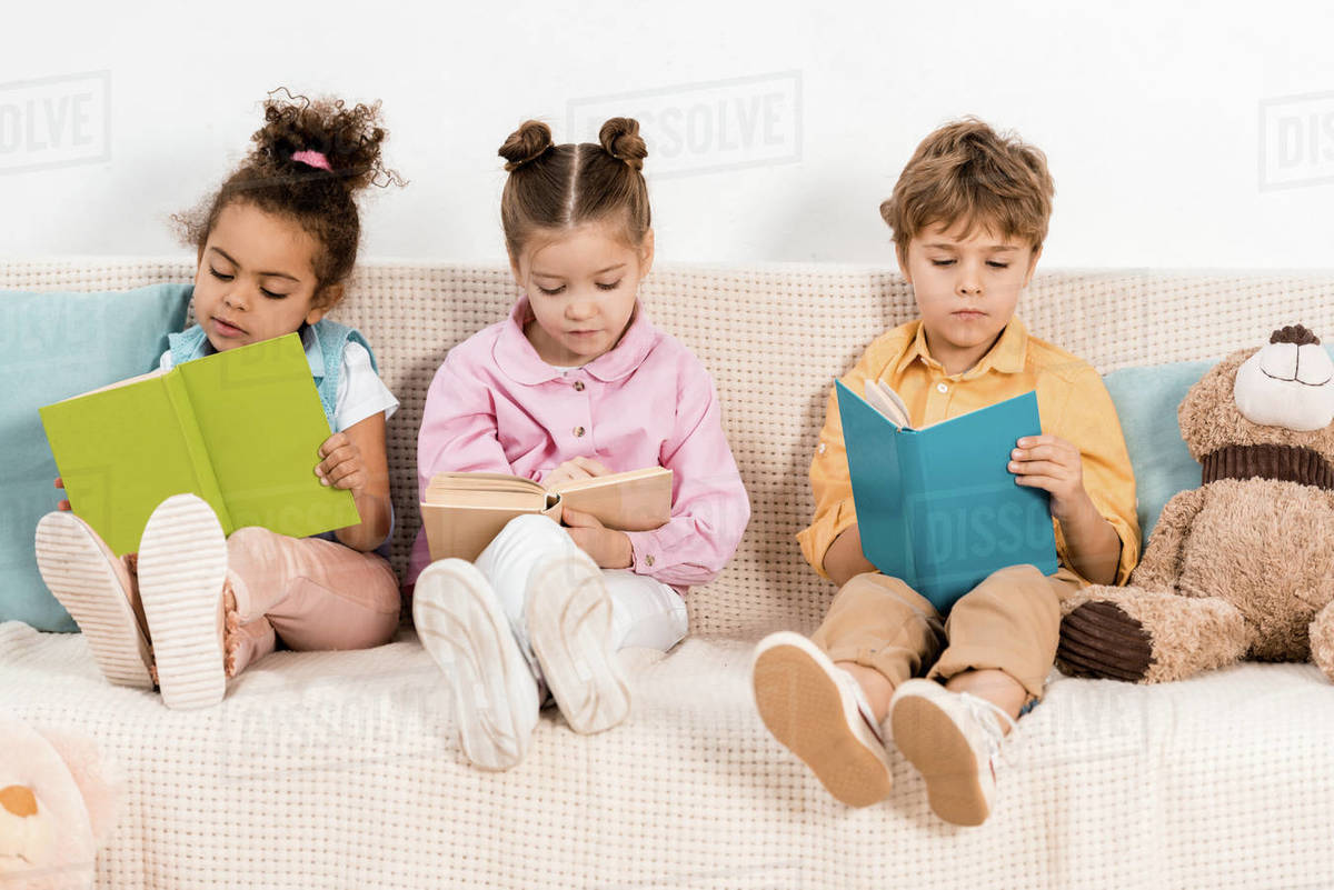 Adorable multiethnic children sitting on sofa and reading books ...